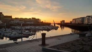 Uma marina com barcos atracados, um calçadão e edifícios ao fundo durante o pôr do sol.