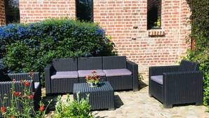 Um edifício de tijolos com um pátio, mobiliado com móveis de vime, e um pequeno jardim com flores.
