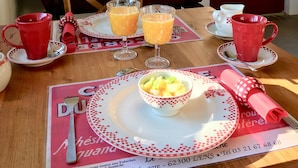 Mesa de pequeno-almoço com fruta, sumo de laranja e copos vermelhos. Há uma obra de arte com tema natural na parede e uma janela com vista para um muro de tijolos.