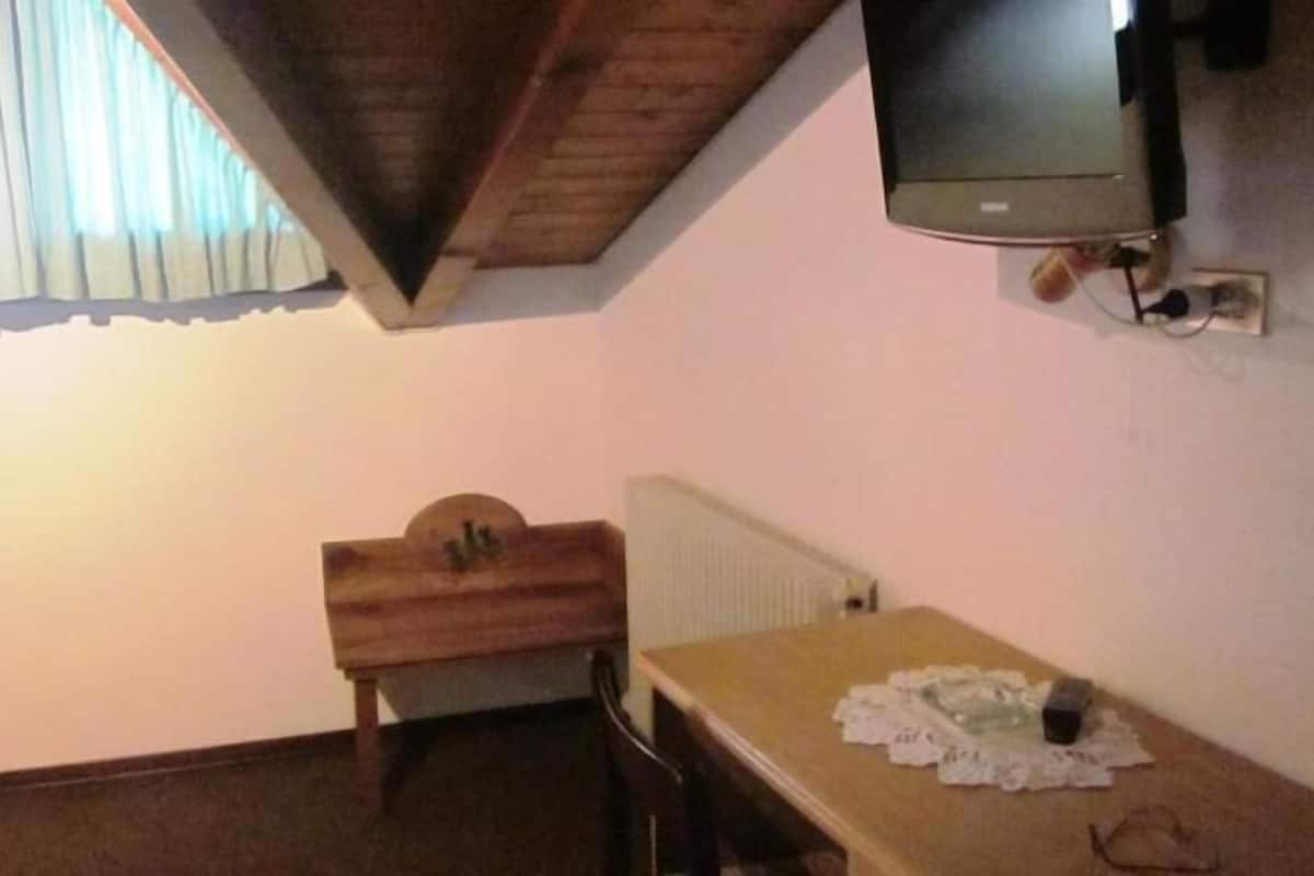 Quarto pequeno com teto de madeira, uma cama de solteiro, mesa de cabeceira com abajur, televisão fixada na parede e janela com cortinas.