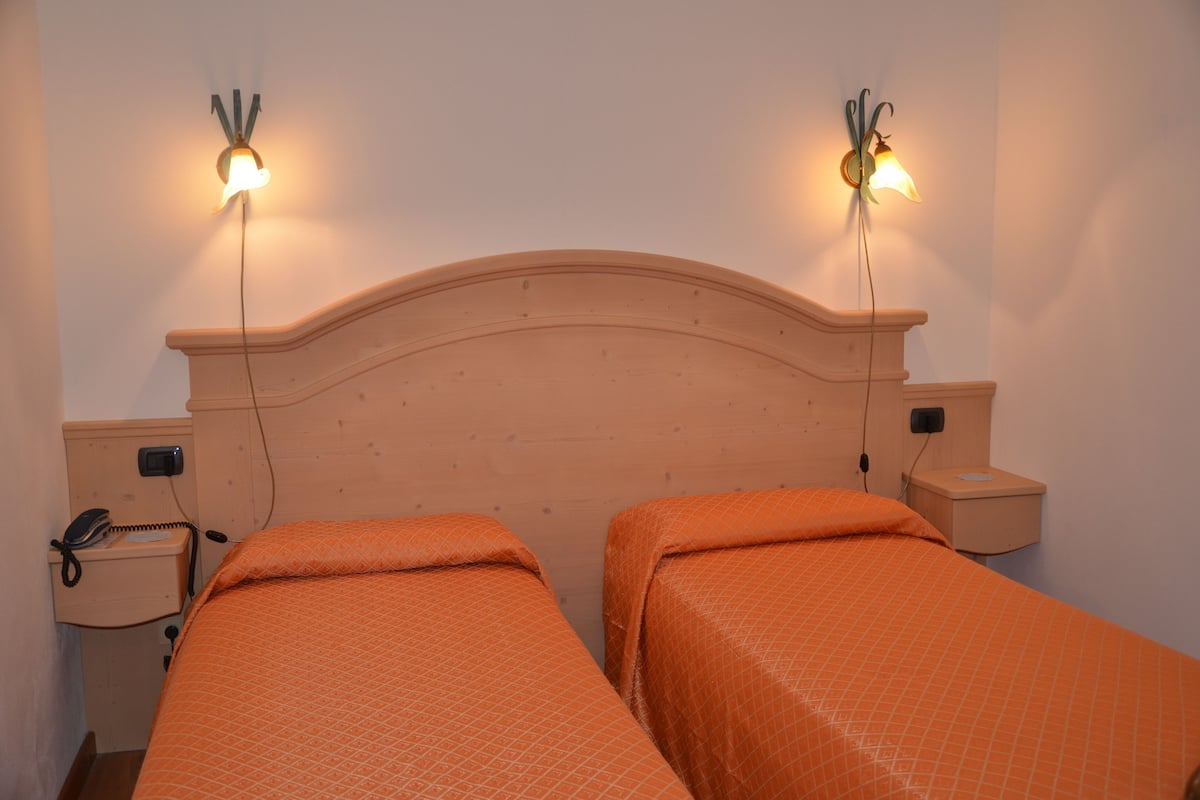 Quarto com duas camas, cada uma com colcha laranja, cabeceira de madeira e luminárias laterais. Em cada lado das camas, tem uma mesa de cabeceira com telefone e luminária.