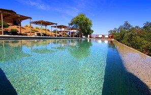 Piscina transparente com espreguiçadeiras e área de estar coberta.
