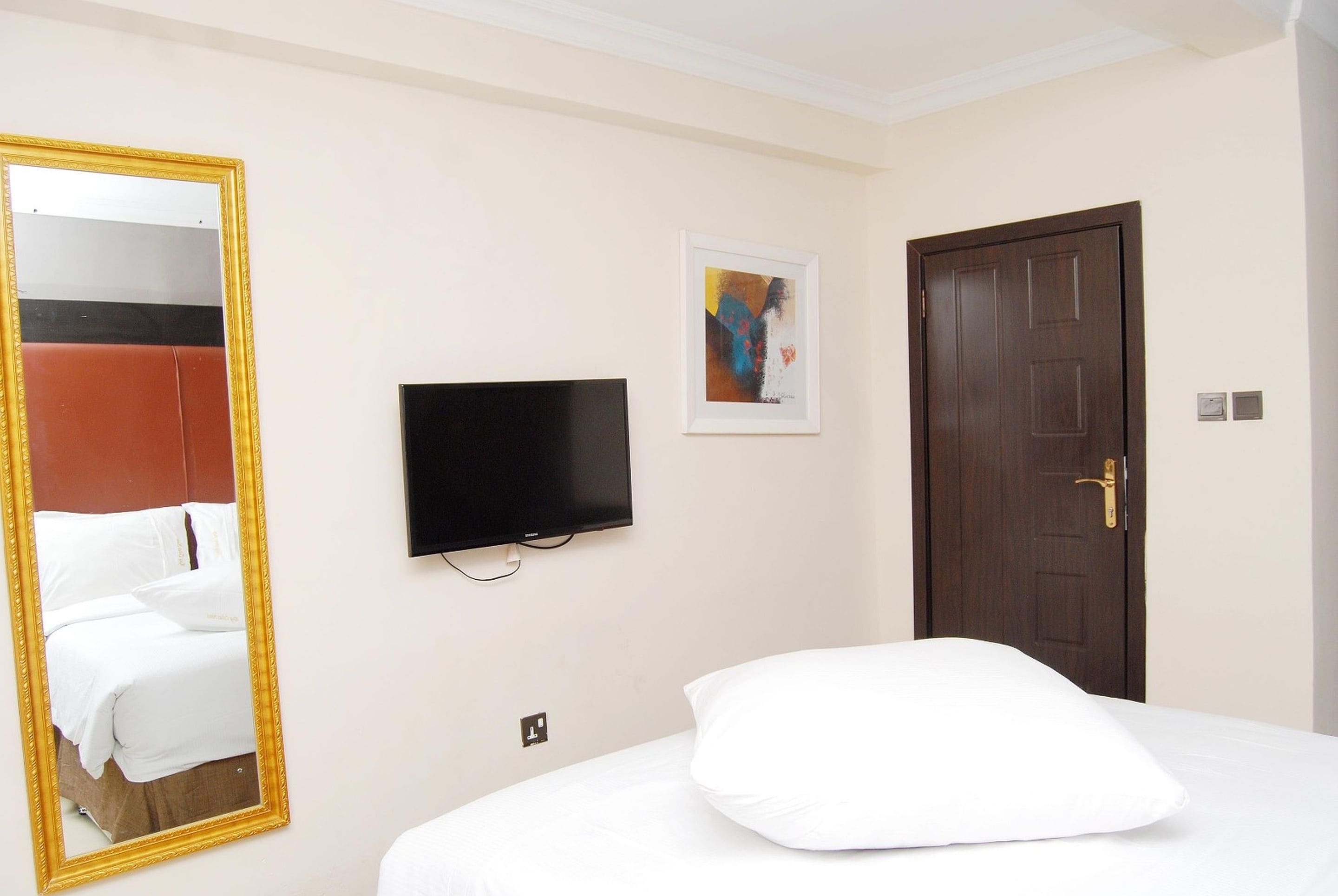 Quarto de hotel com cama, um espelho grande, televisão de tela plana, uma porta e um quadro na parede.