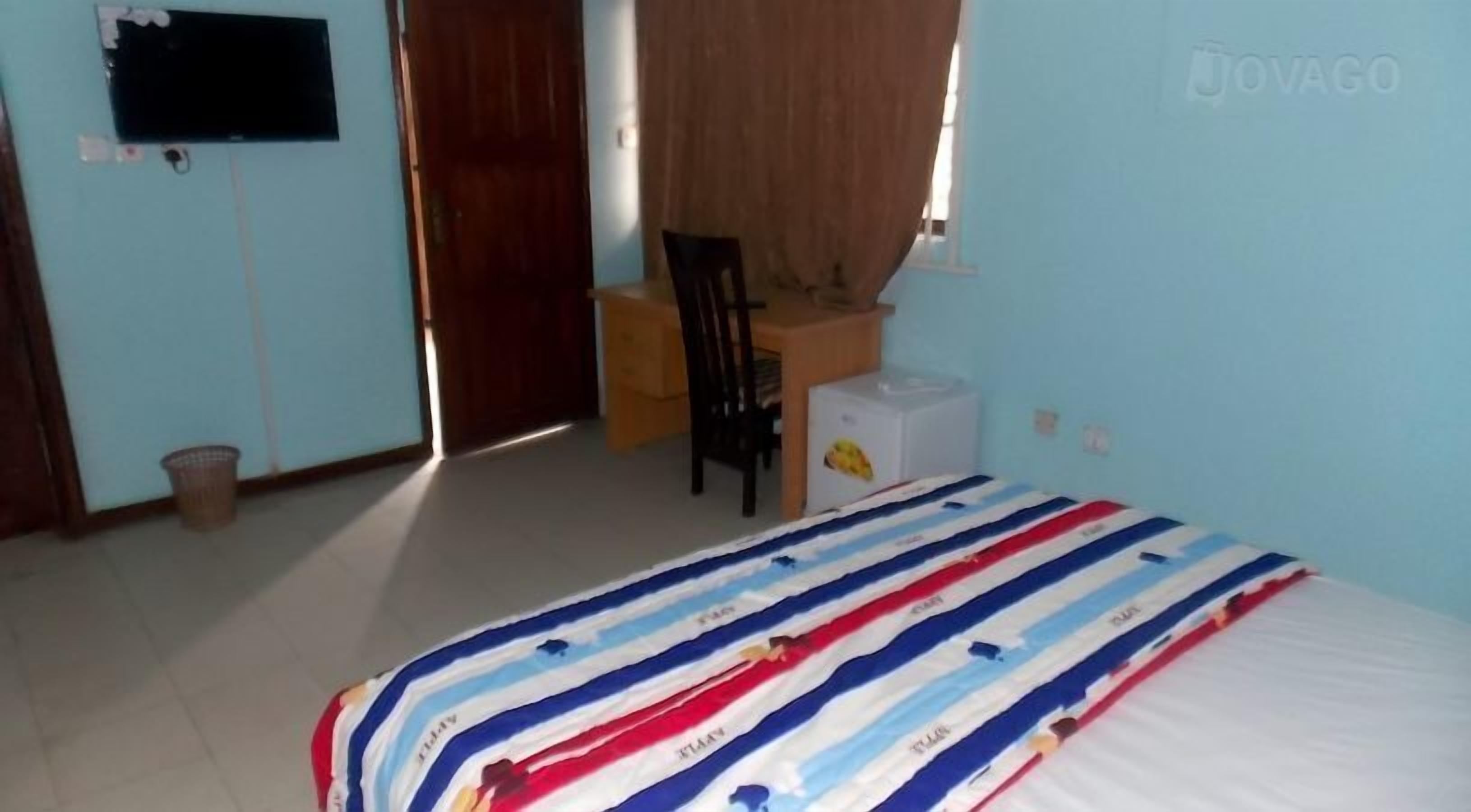 Quarto com cama, uma cadeira de madeira, uma mesinha e uma televisão na parede.