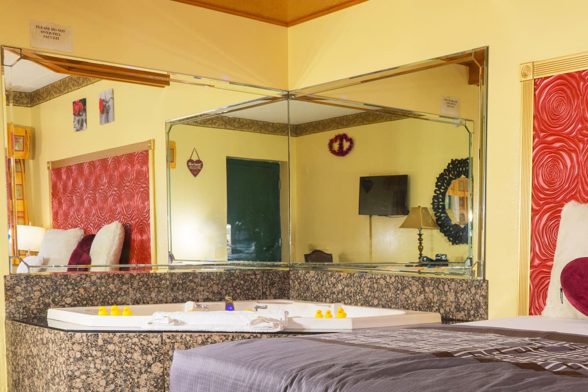 Quarto de hotel com um espelho grande, uma cama com cabeceira estampada em vermelho e branco, uma toucador com lavatório e um design floral vermelho na parede.