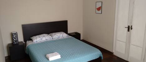 Cama bem arrumada com colcha azul, duas mesas de cabeceira, uma delas com controle remoto, e uma porta à direita.
