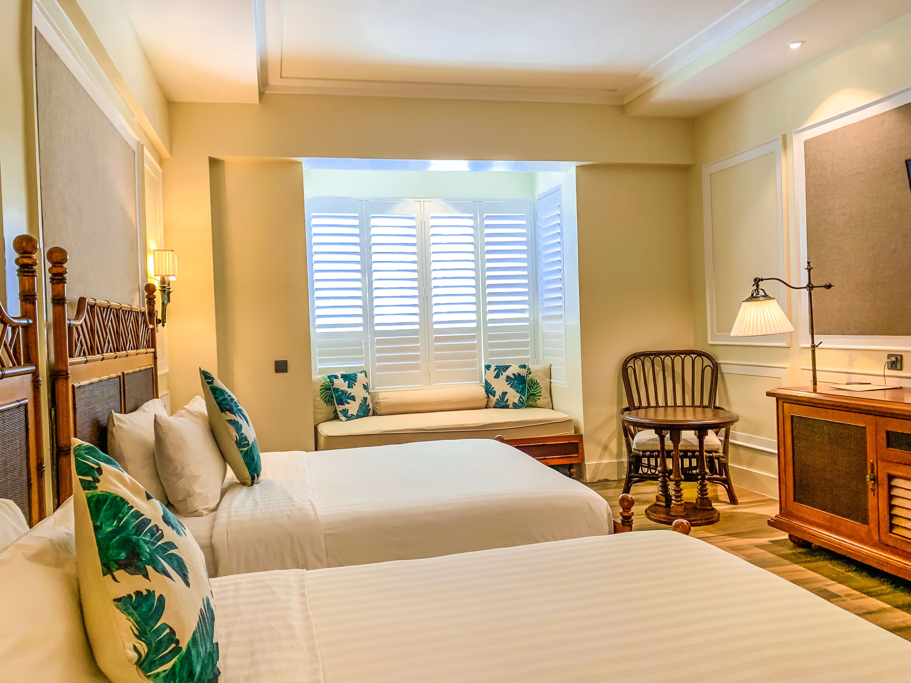 Quarto de hotel com duas camas, uma janela com persianas, uma cadeira, uma escrivaninha e um abajur.