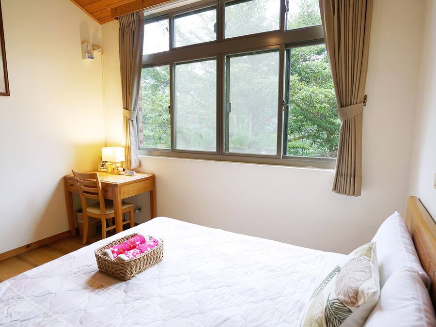 Quarto com cama, escrivaninha com cadeira, janela com cortinas e televisão fixada na parede.