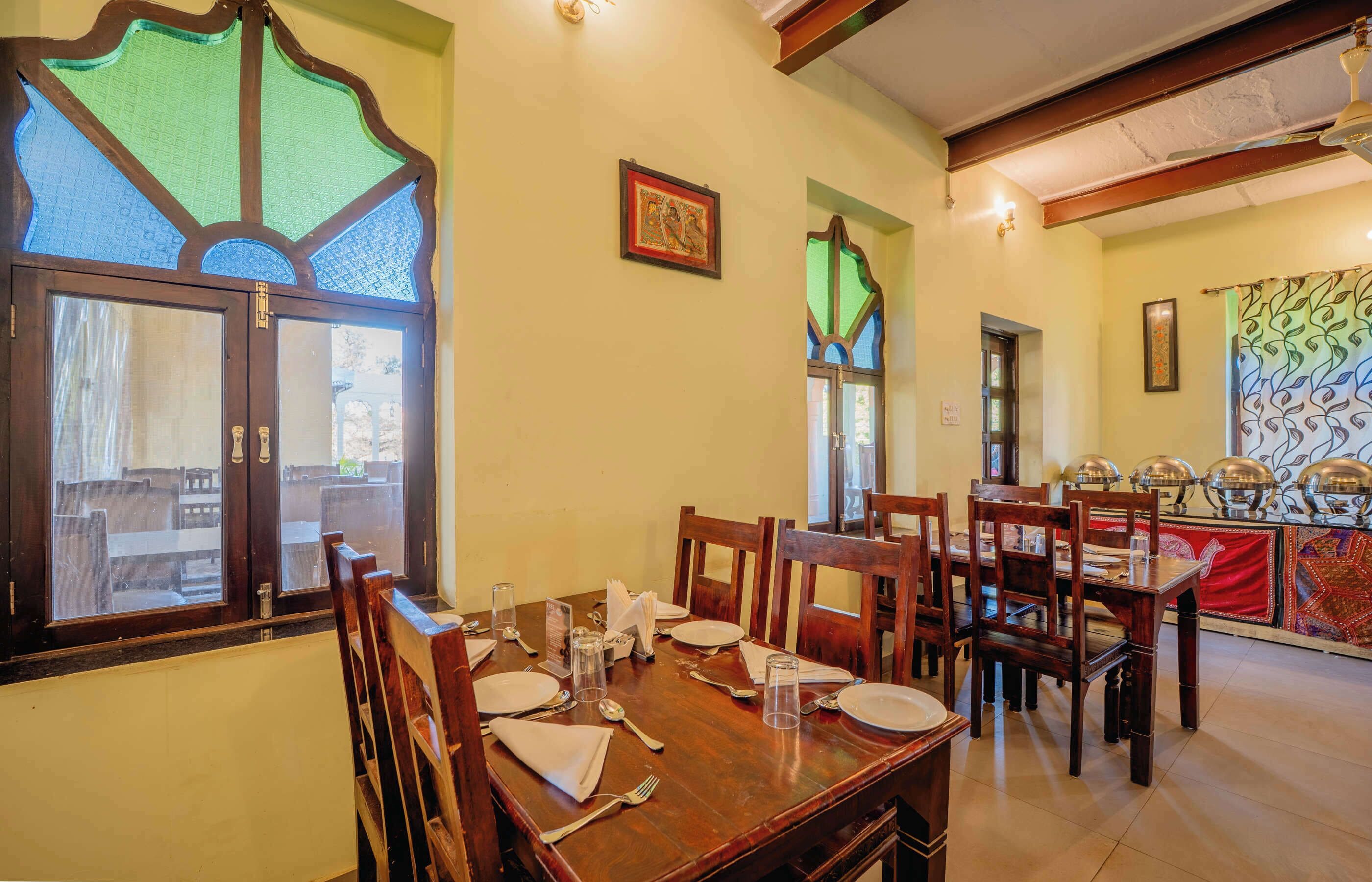 Uma sala de jantar com mesas e cadeiras de madeira, um buffet montado e painéis de janela decorativos.
