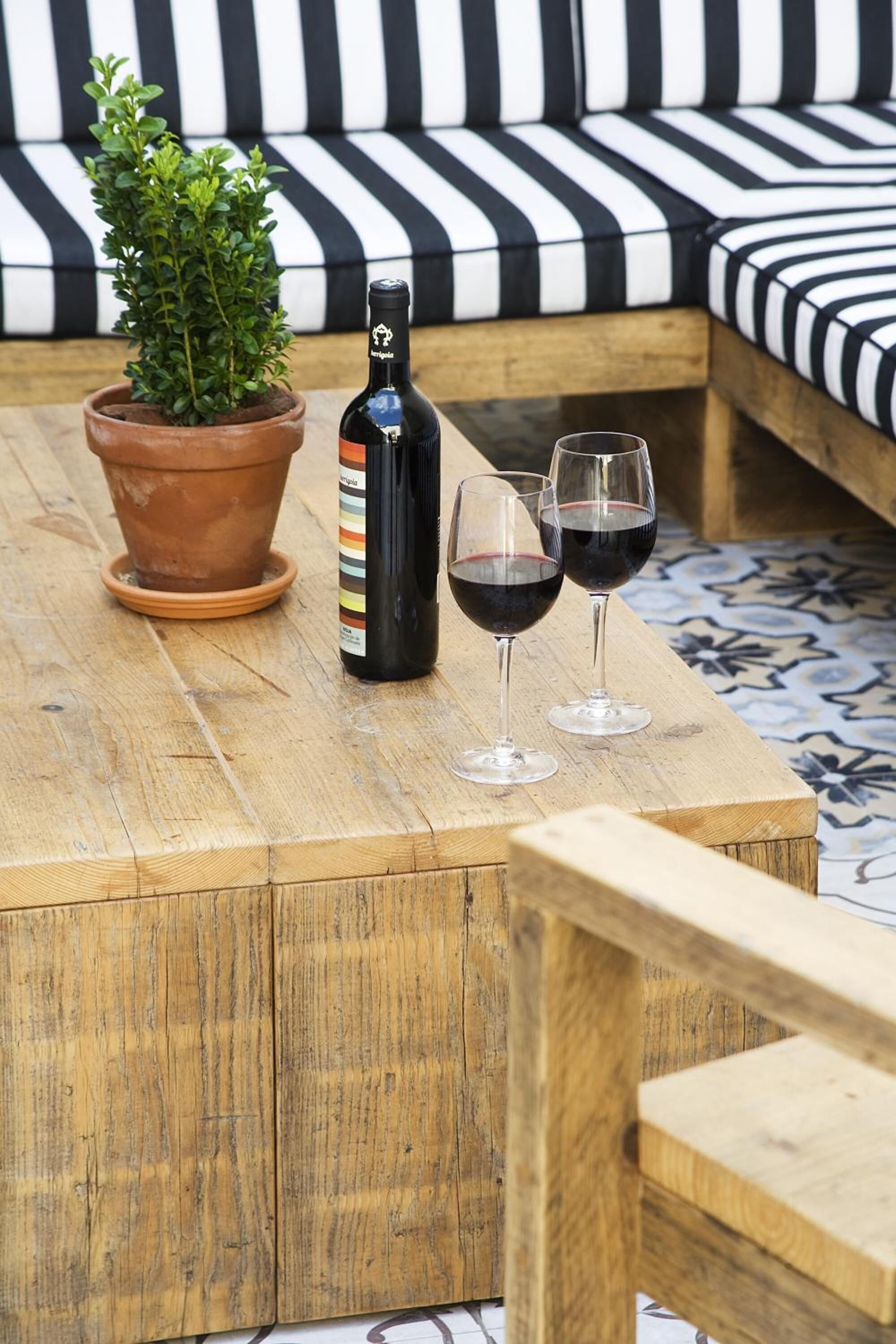 Mesa de madeira com uma garrafa e duas taças de vinho, uma planta em vaso e um sofá listrado ao fundo.