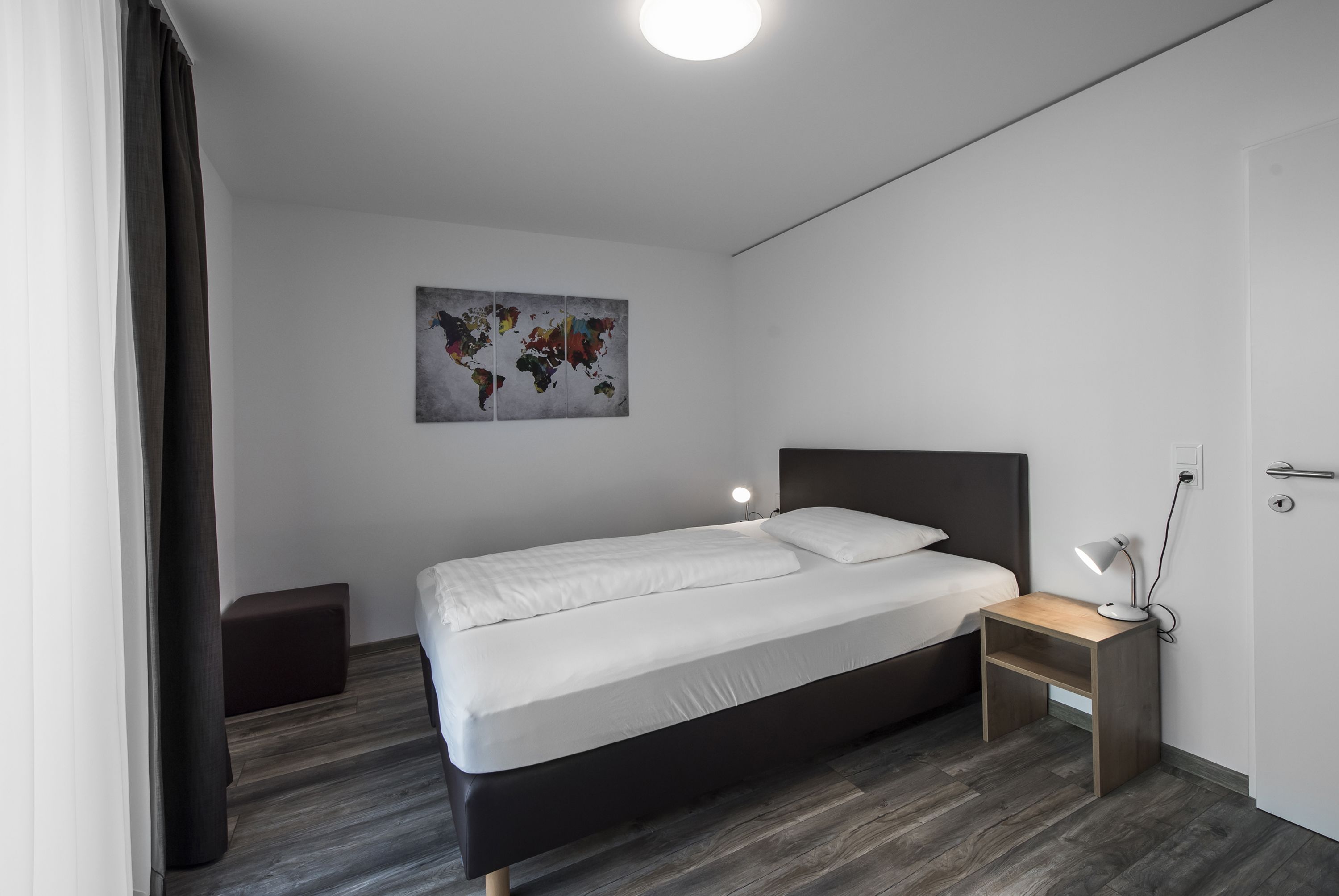 Quarto de hotel moderno com uma cama de solteiro, mesa de cabeceira com abajur, uma obra de arte com um mapa-múndi na parede e chão de madeira escura.