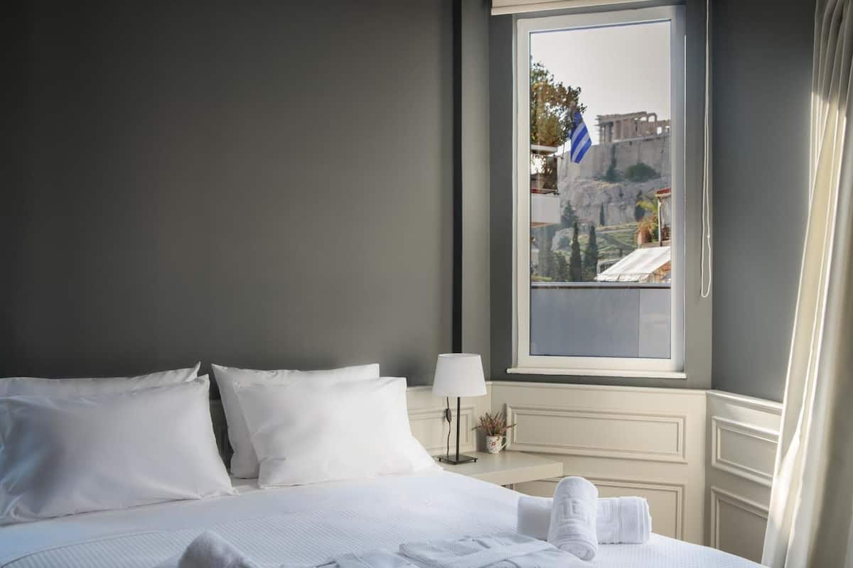 Quarto de hotel com uma cama grande, uma mesa de cabeceira, uma luminária e uma janela com vista para um edifício histórico.