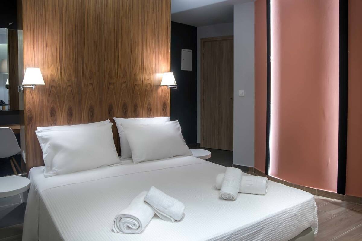 Quarto de hotel com cabeceira de madeira, cama com roupa de cama branca, duas luminárias de apoio e cama bem arrumada com toalhas dobradas.