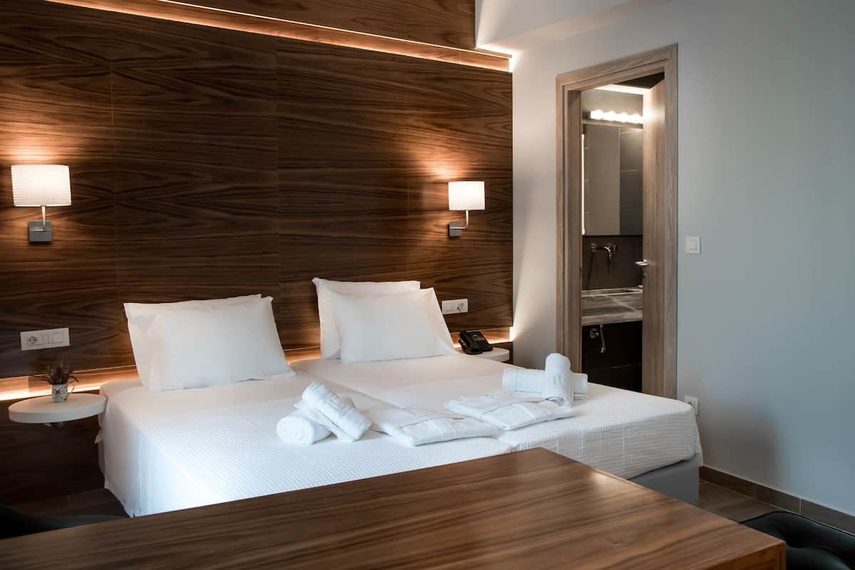Quarto de hotel com cabeceira de madeira, cama com roupa de cama branca, duas luminárias de cabeceira, mesa de cabeceira de madeira e banheiro visível através de uma porta aberta.