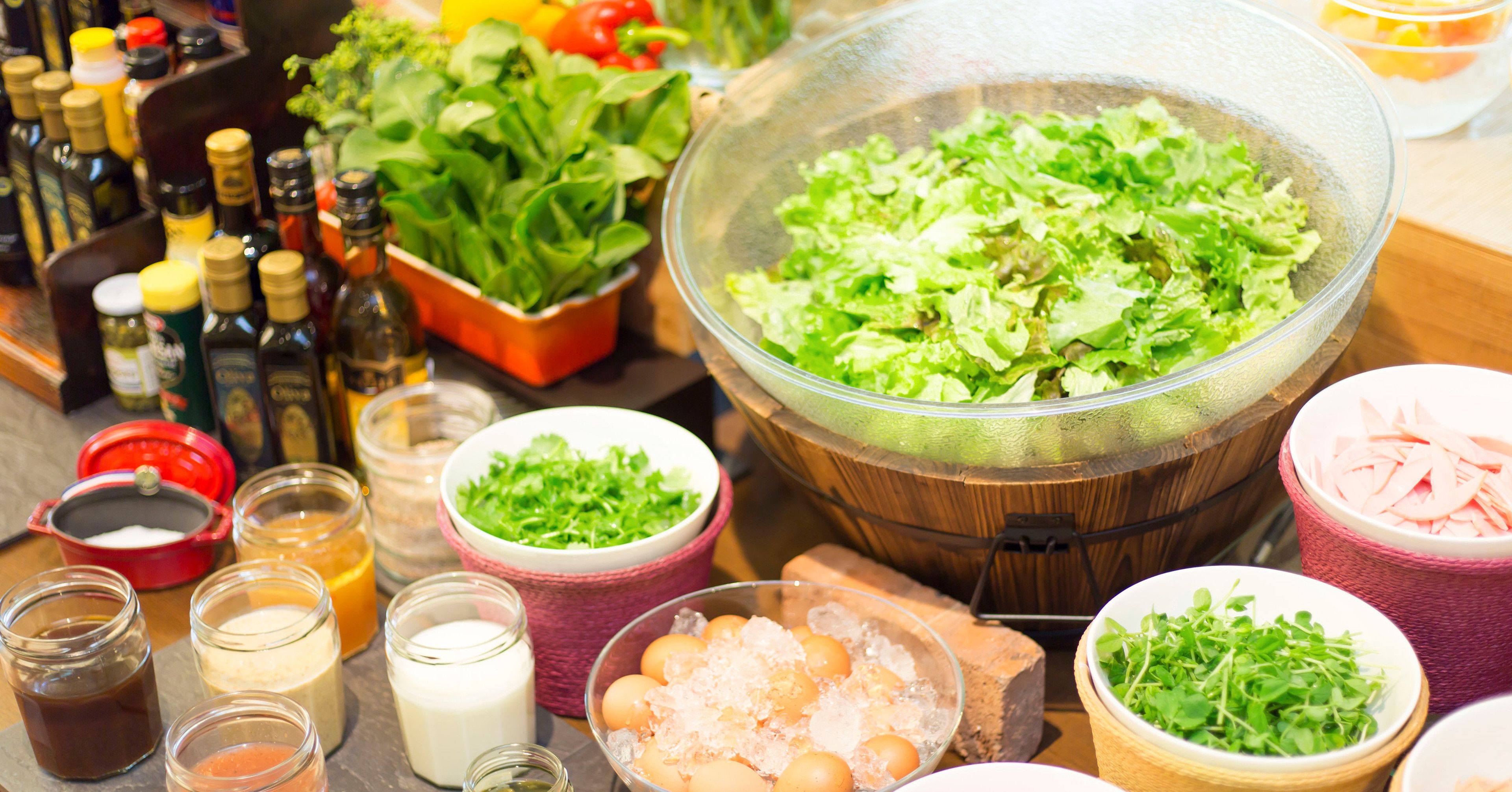 Diversos alimentos e acompanhamentos em uma mesa, incluindo uma grande salada, potes com condimentos e tigelas com vegetais e ovos.