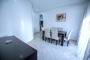 Uma área de refeições com uma mesa de madeira escura e cadeiras brancas, um quadro na parede e uma cozinha ao fundo.