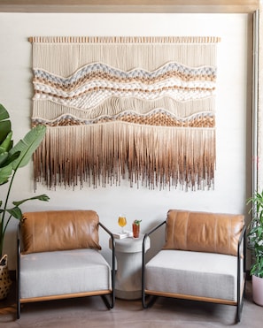 Um quarto com duas poltronas de couro, uma mesinha redonda com bebidas e um painel de macramé na parede.