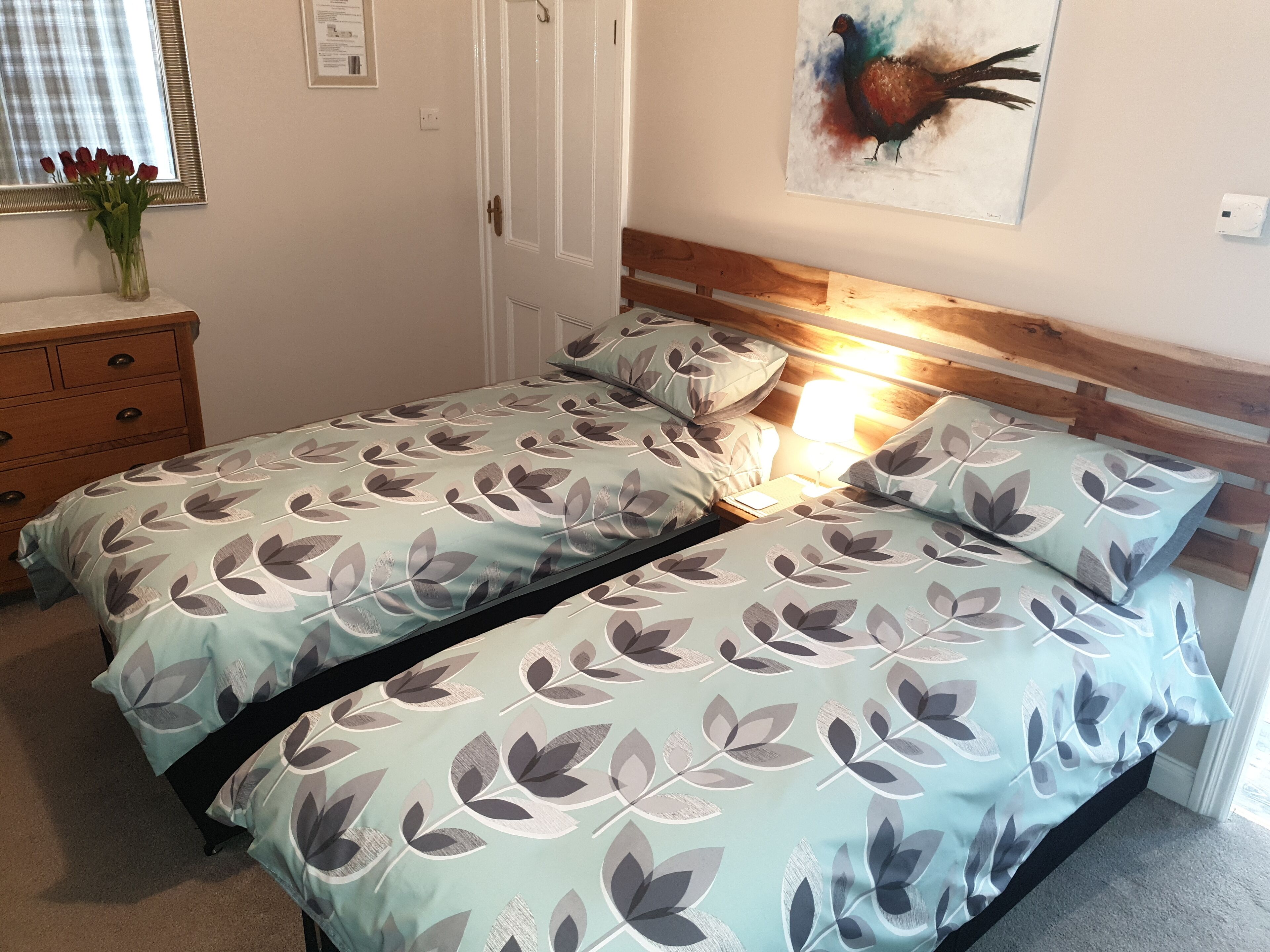 Quarto com cama estampada com padrão de folha, cabeceira de madeira, mesa de cabeceira, luminária e um quadro na parede.