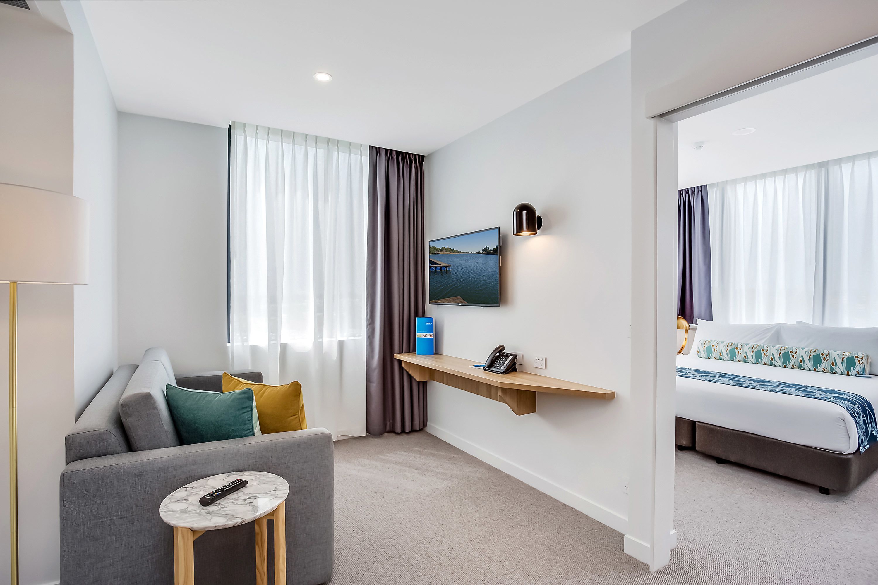 Quarto de hotel moderno com sofá cinzento, mesa redonda pequena com um comando, cama com colcha estampada, TV fixada na parede e telefone também na parede.