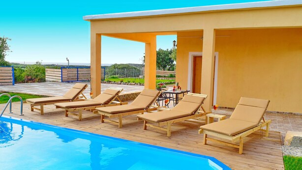 Uma área junto à piscina com espreguiçadeiras e uma mesa, rodeada por um deck de madeira e um edifício com fachada bege.