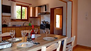 Uma sala de jantar com uma mesa de madeira posta para quatro, uma cozinha ao fundo e uma janela com cortina.
