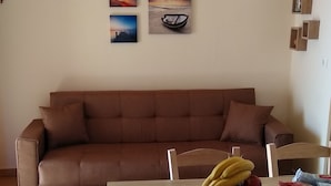Uma sala de estar com um sofá marrom, uma mesa de jantar de madeira com uma tigela de frutas e duas imagens penduradas na parede.