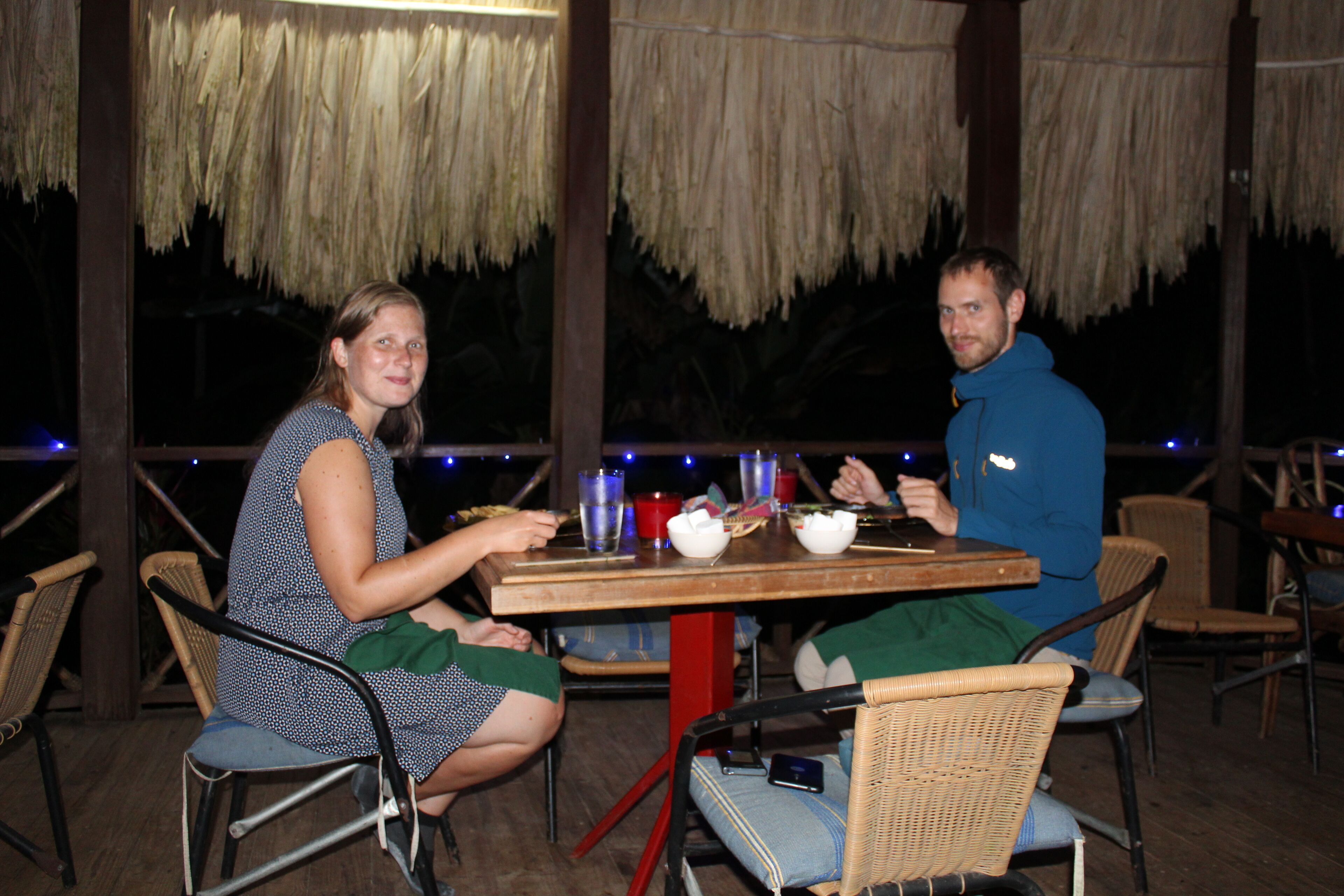 Um casal jantando em restaurante ao ar livre com teto de palha e cadeiras de vime.