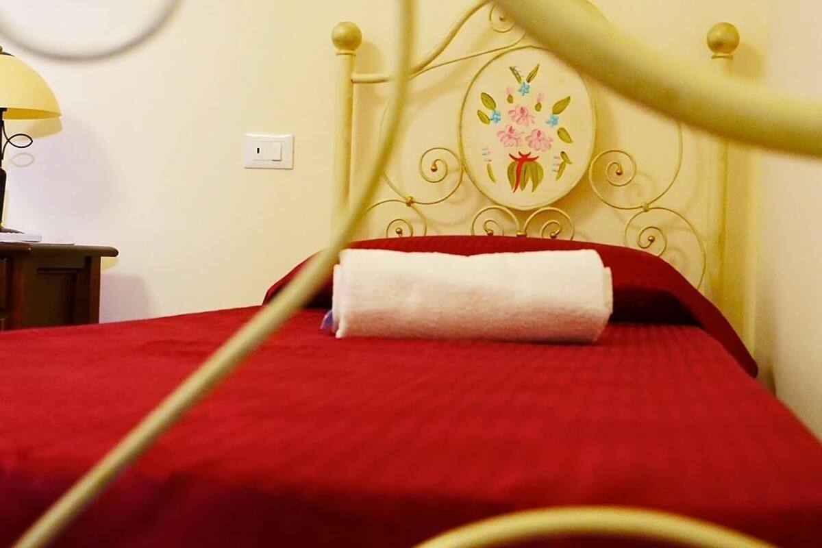 Uma cama com um cobreleito vermelho e uma toalha branca, um abajur em uma mesa de cabeceira e uma parede com um prato decorativo.