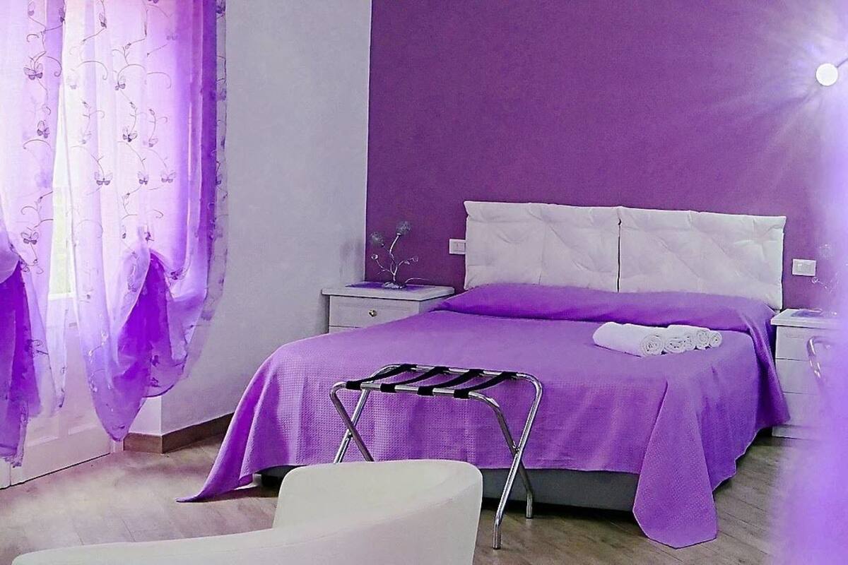 Quarto com colcha roxa, almofadas brancas, mesa de cabeceira pequena com abajur e uma cadeira.
