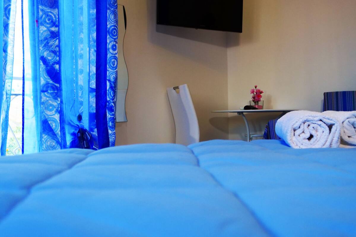 Quarto de hotel com uma cama, uma mesa pequena com um vaso de flores e uma televisão na parede.