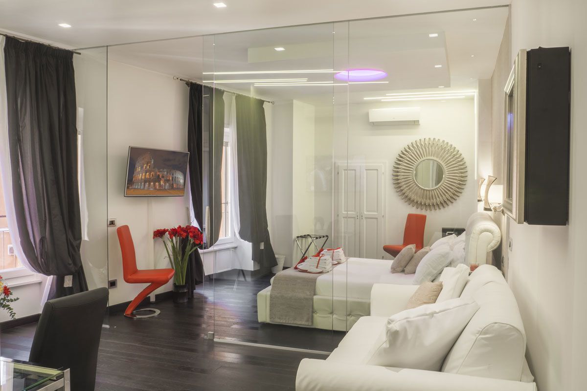 Quarto de hotel moderno com divisória de vidro, sofá branco, cadeira vermelha e um quadro de um edifício.