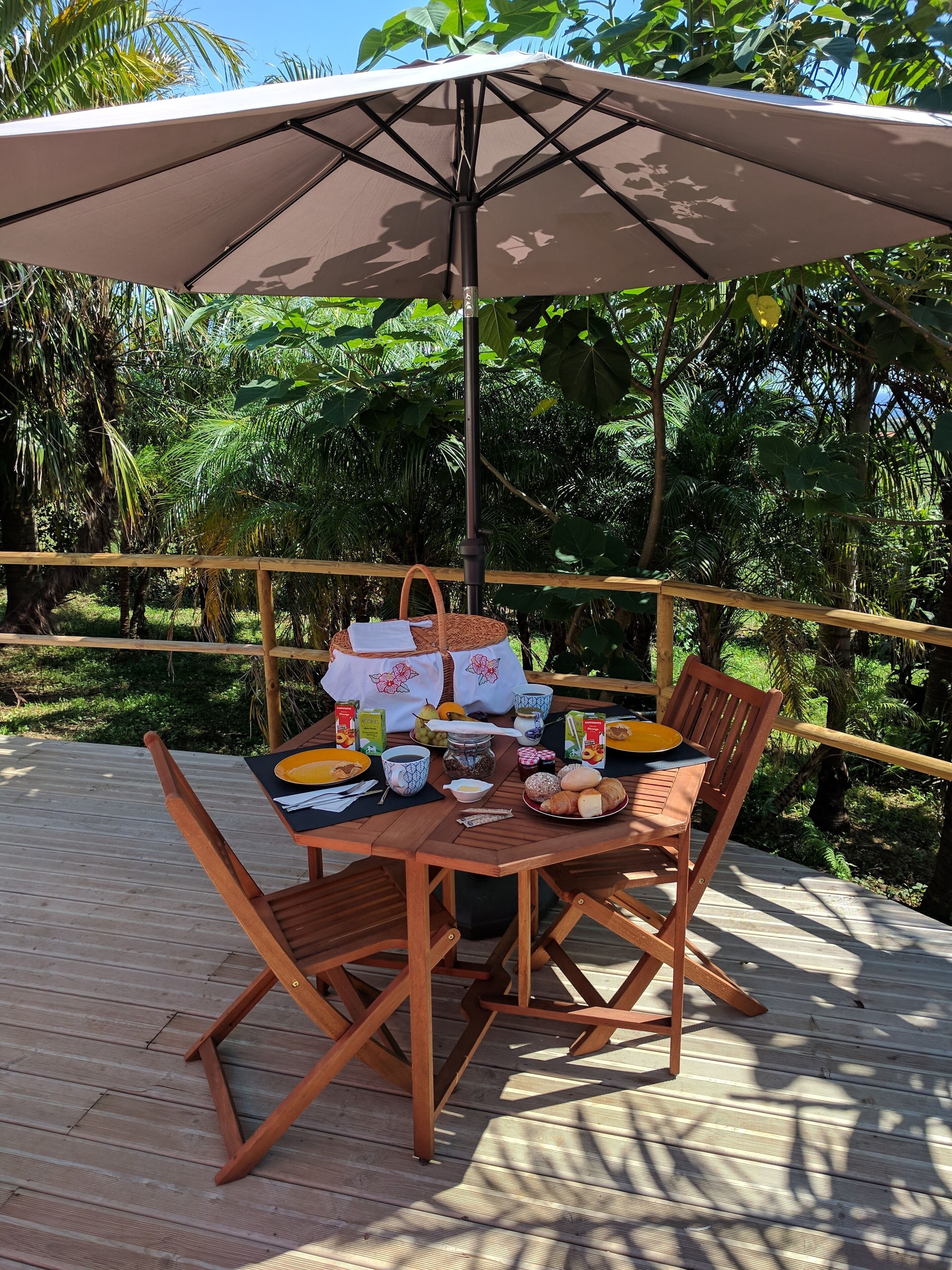 Um deck de madeira com uma mesa posta para o café da manhã, rodeado por vegetação exuberante.