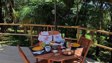 Um deck de madeira com uma mesa posta para o café da manhã, rodeado por vegetação exuberante.