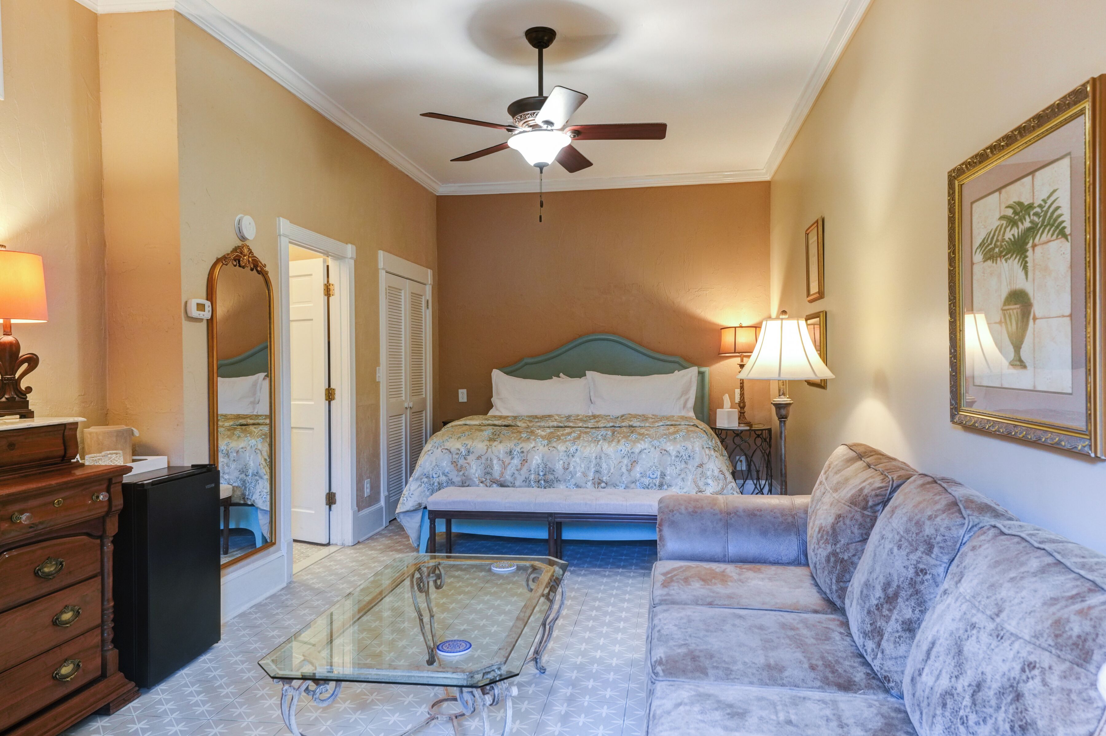 Quarto com cama, sofá, mesa de centro de vidro, cômoda, ventilador de teto e uma porta.