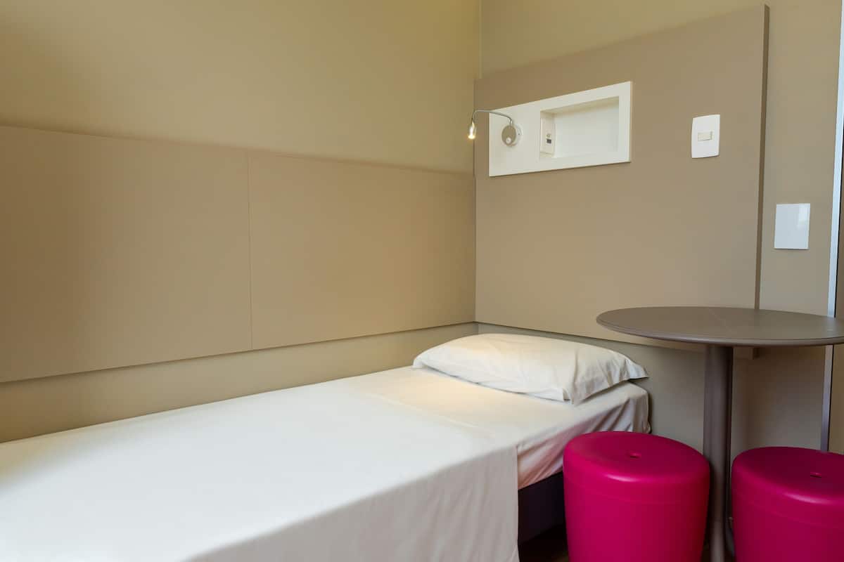 Uma cama de solteiro com colchão e travesseiro brancos, uma mesa pequena com tampo redondo e dois banquinhos rosa.