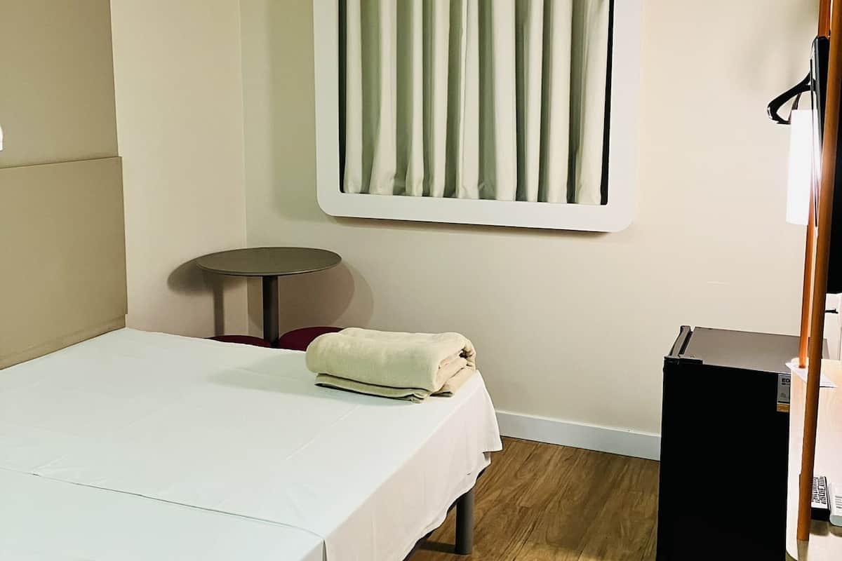 Quarto pequeno e bem organizado, com uma cama, uma mesinha, janela com cortinas e um frigorífico compacto.