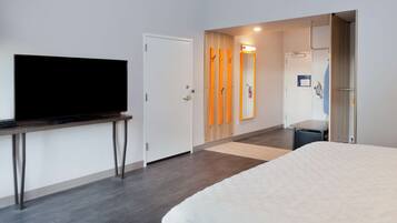 Quarto de hotel moderno com uma cama grande, televisão de tela plana em um suporte, porta de madeira com espelho e área de chuveiro.