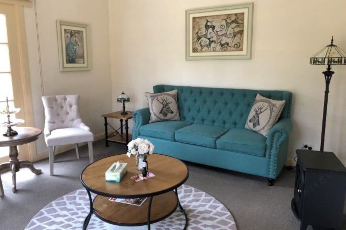 Uma sala de estar com um sofá verde-água, uma poltrona branca, uma mesa de centro redonda com um vaso de flores e um quadro na parede.