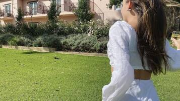 Uma pessoa vestida de branco está em um gramado com uma casa ao fundo.