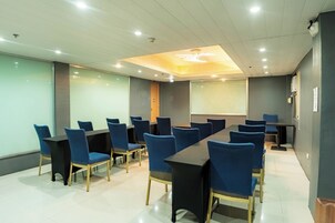 Sala de conferências com uma longa mesa, cadeiras azuis, tela de projetor e teto branco com iluminação embutida.