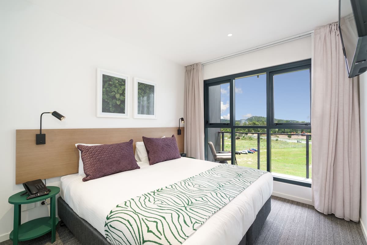 Quarto de hotel com uma cama grande, uma mesinha de cabeceira, uma mesa lateral verde, uma janela com vista para o exterior e duas quadros na parede.