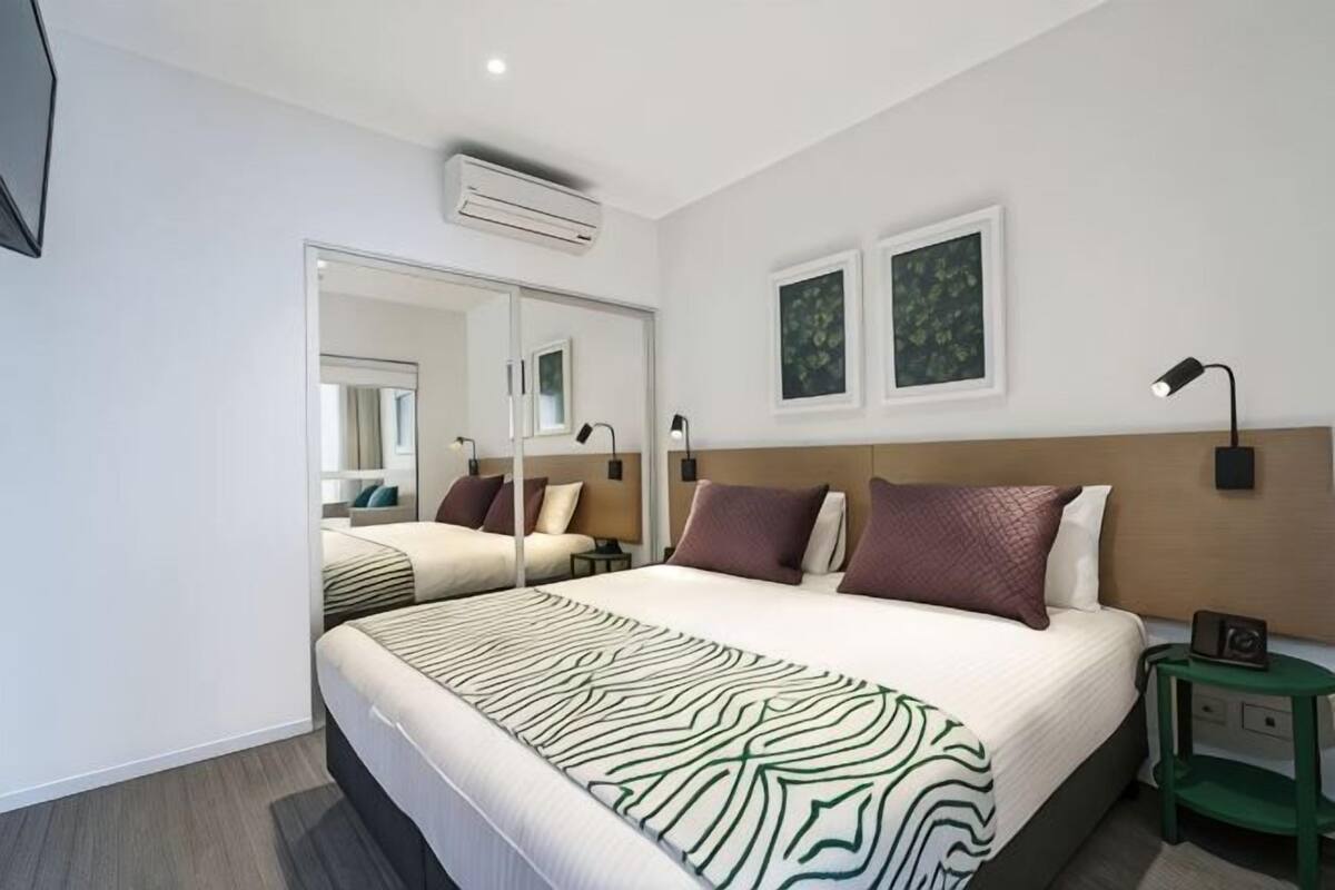 Quarto de hotel com uma cama grande, uma mesinha de cabeceira, um espelho e duas fotos emolduradas na parede.