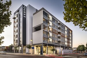 Um hotel com vários andares, um destaque para o letreiro 'QUEST', grandes janelas e um design arquitetônico moderno.