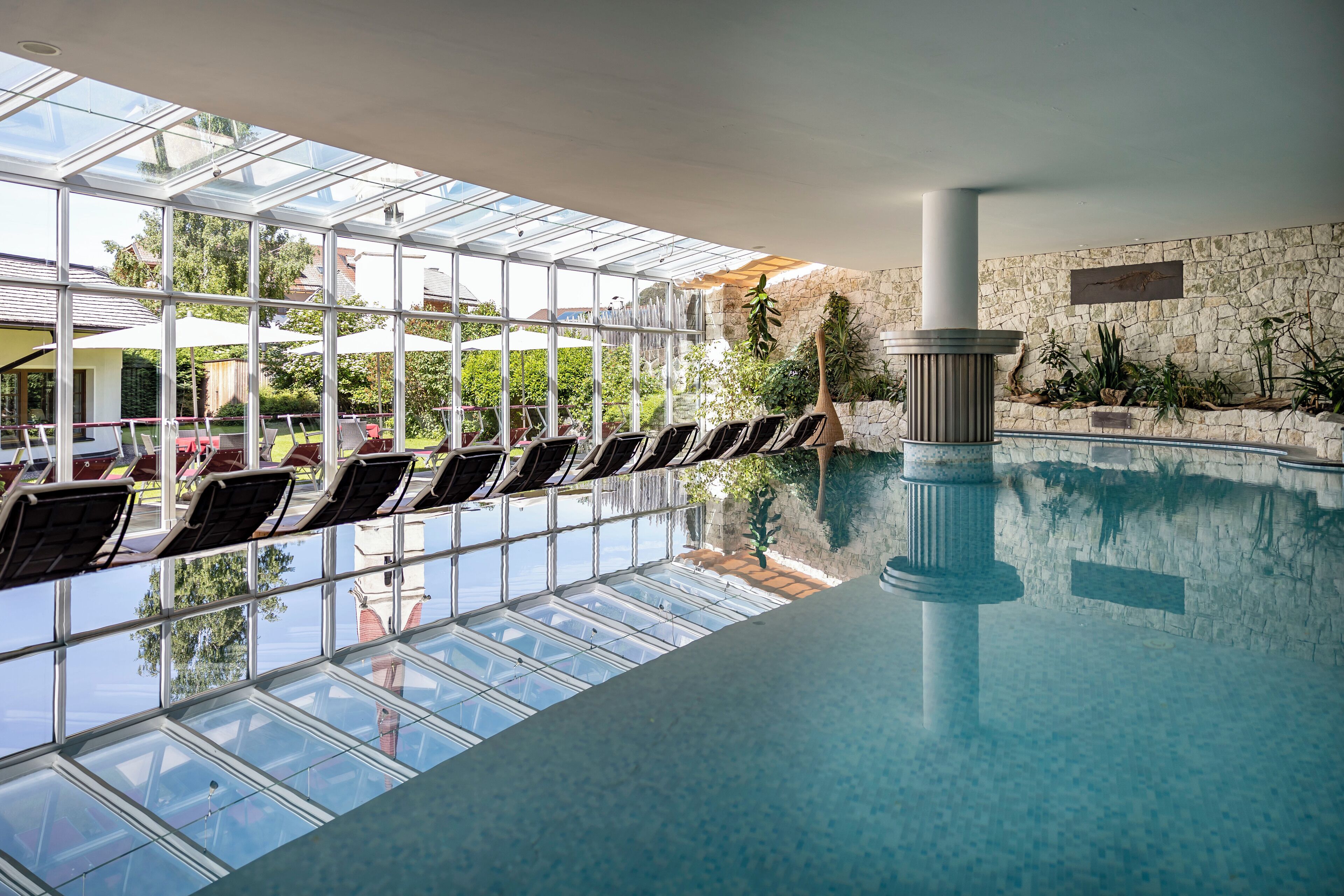 Uma grande piscina interna com teto de vidro, rodeada por espreguiçadeiras e plantas em vasos.