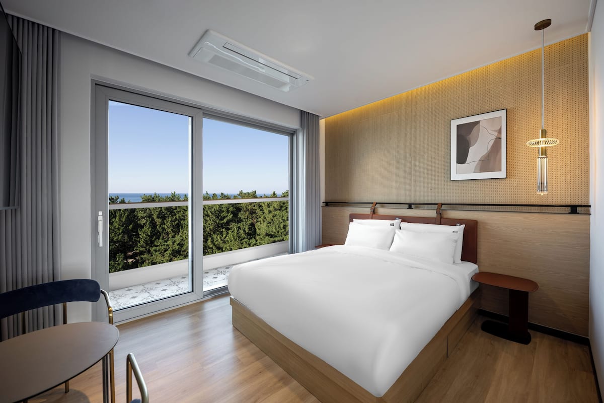 Quarto de hotel moderno com uma cama grande, uma escrivaninha e uma cadeira. Há uma janela ampla com vista para o exterior.