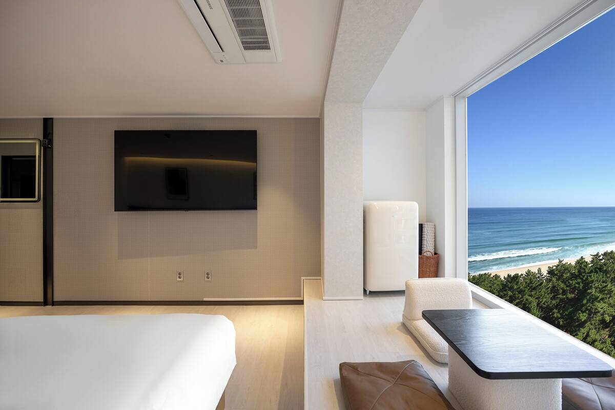 Quarto de hotel moderno com uma cama grande, televisão de tela plana na parede, área de estar com mesa e vista para o oceano.