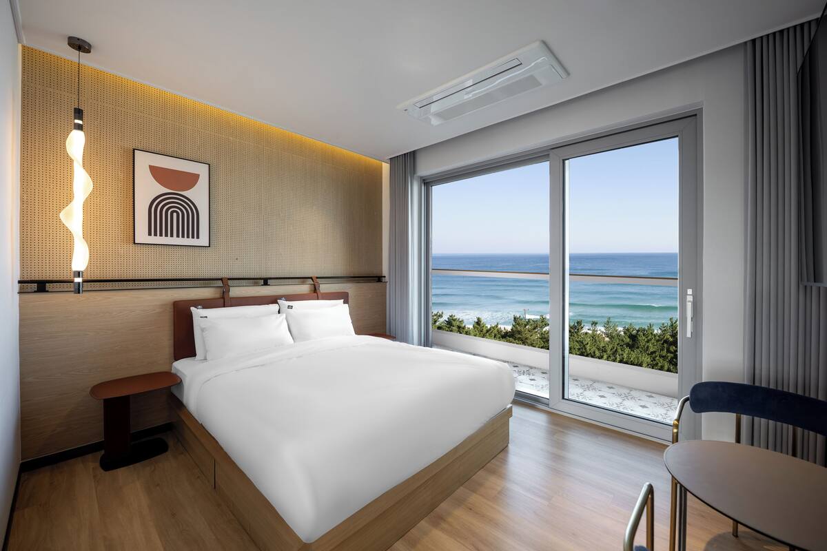 Quarto de hotel moderno com uma cama grande, mesa de cabeceira, uma cadeira e vista para o oceano.