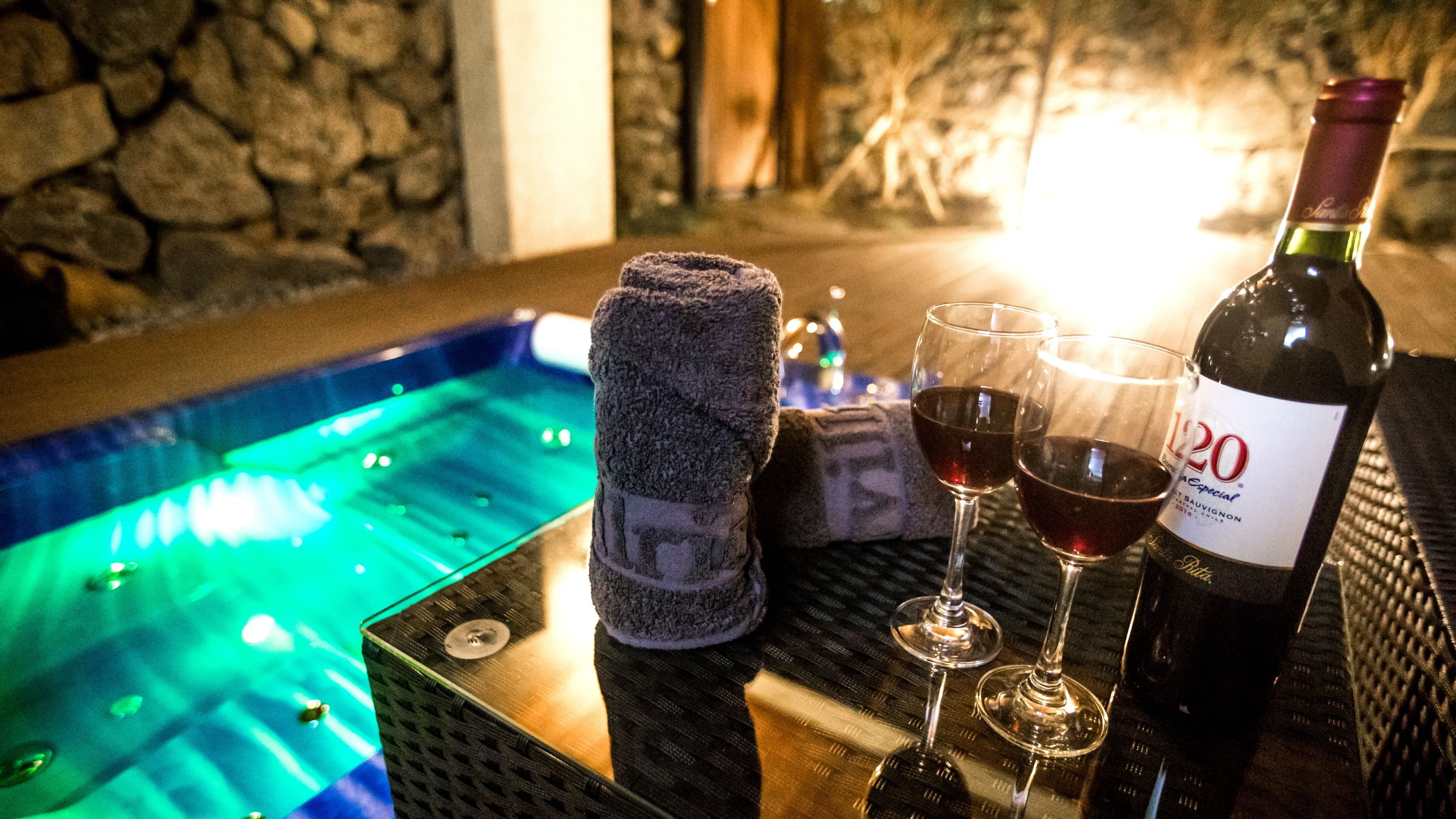 Uma banheira de hidromassagem com parede de pedra ao fundo, dois copos de vinho e uma garrafa sobre uma mesa.