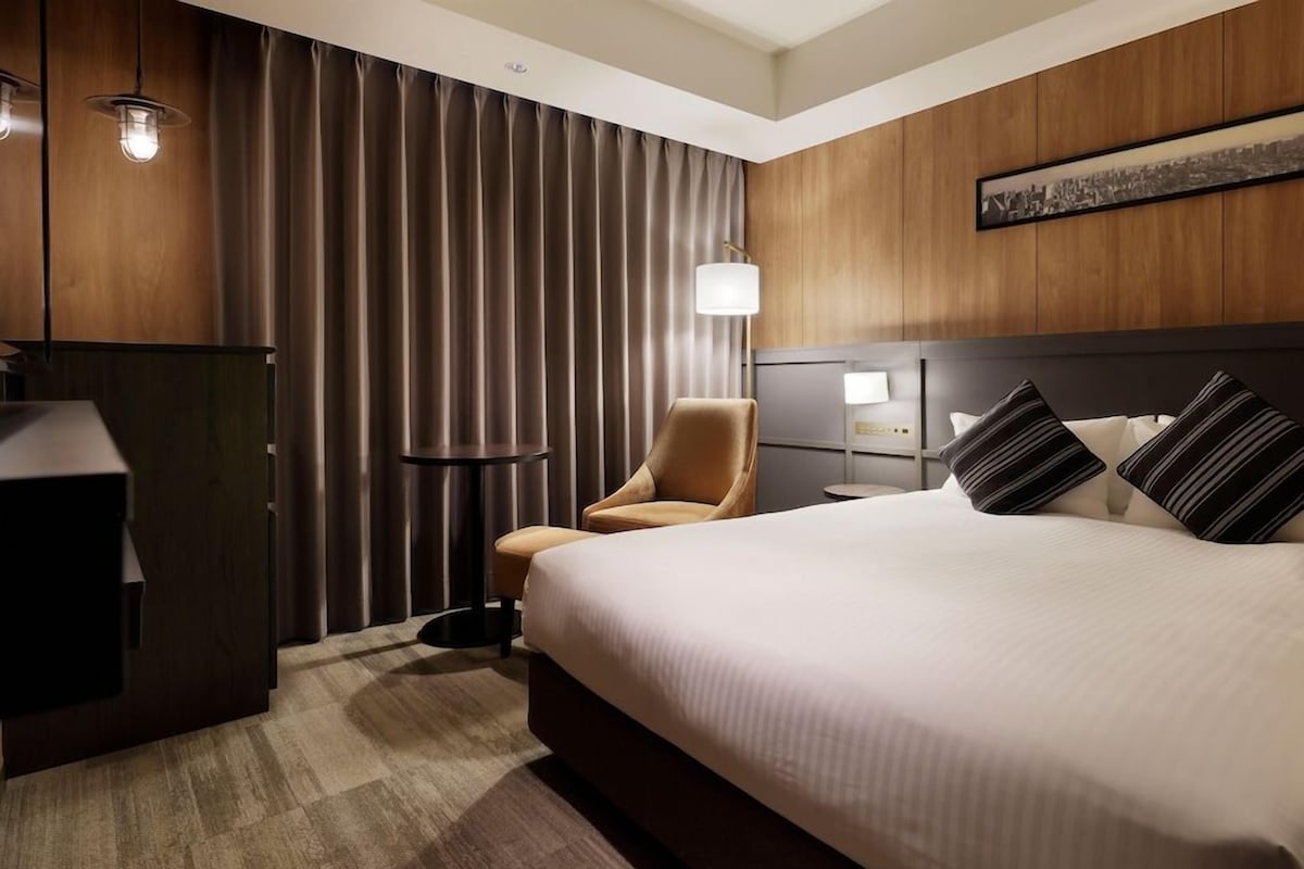 Quarto de hotel com uma cama grande, uma cadeira, uma escrivaninha, um abajur e uma fotografia de paisagem urbana na parede.