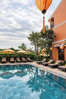 Área de piscina com espreguiçadeiras e guarda-sóis, cercada por vegetação e um edifício com varanda.